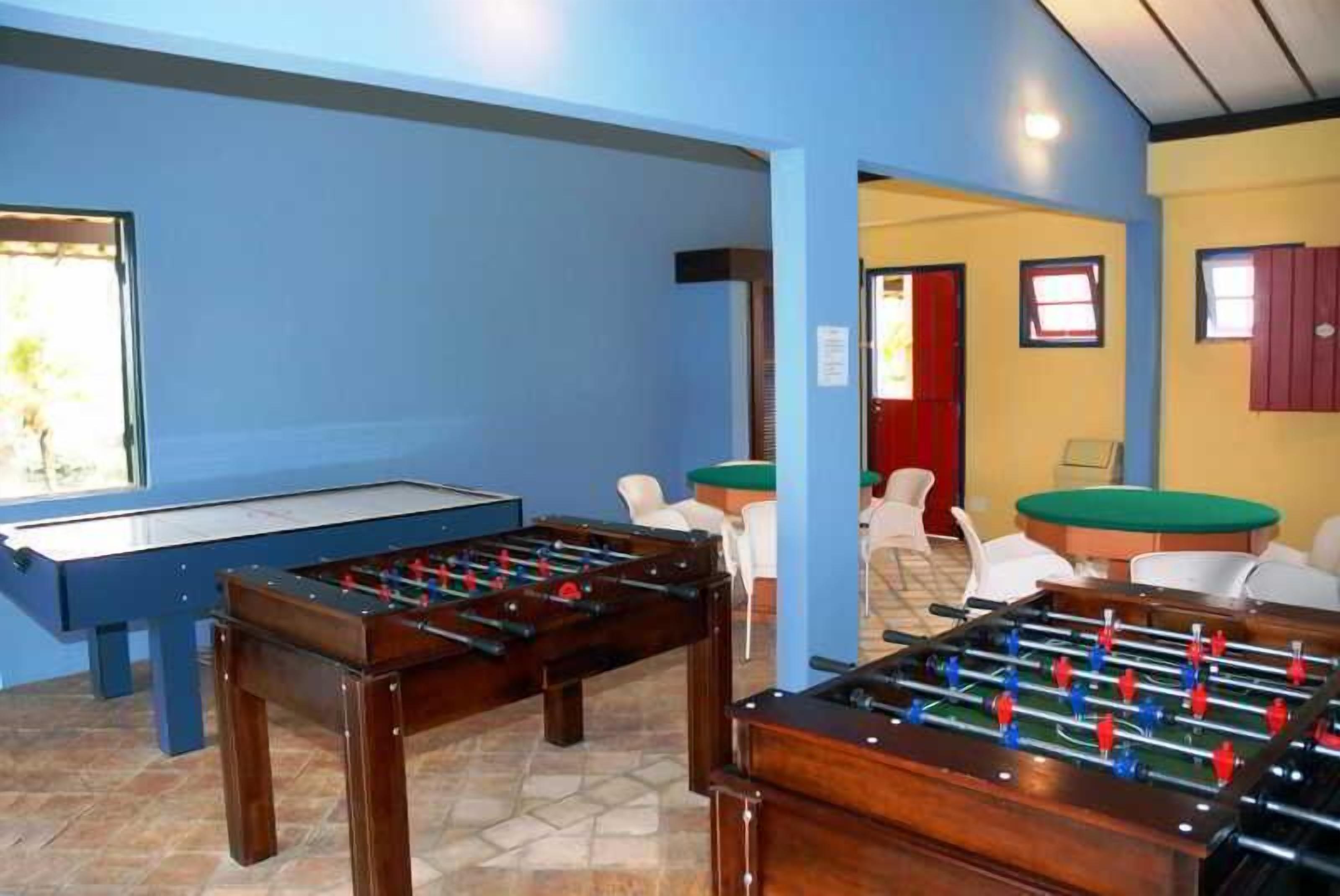 Quarto com duas mesas de futebol de mesa, uma mesa de bilhar e uma mesa verde, com cadeiras e uma porta ao fundo.