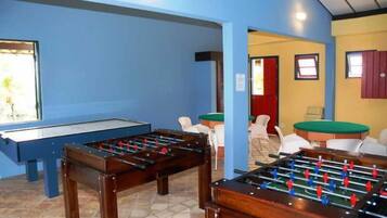 Quarto com duas mesas de futebol de mesa, uma mesa de bilhar e uma mesa verde, com cadeiras e uma porta ao fundo.