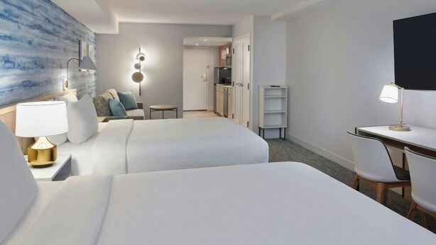 Quarto de hotel com duas camas, uma escrivaninha, uma cadeira, uma luminária, TV fixada na parede e uma cozinha compacta ao fundo.