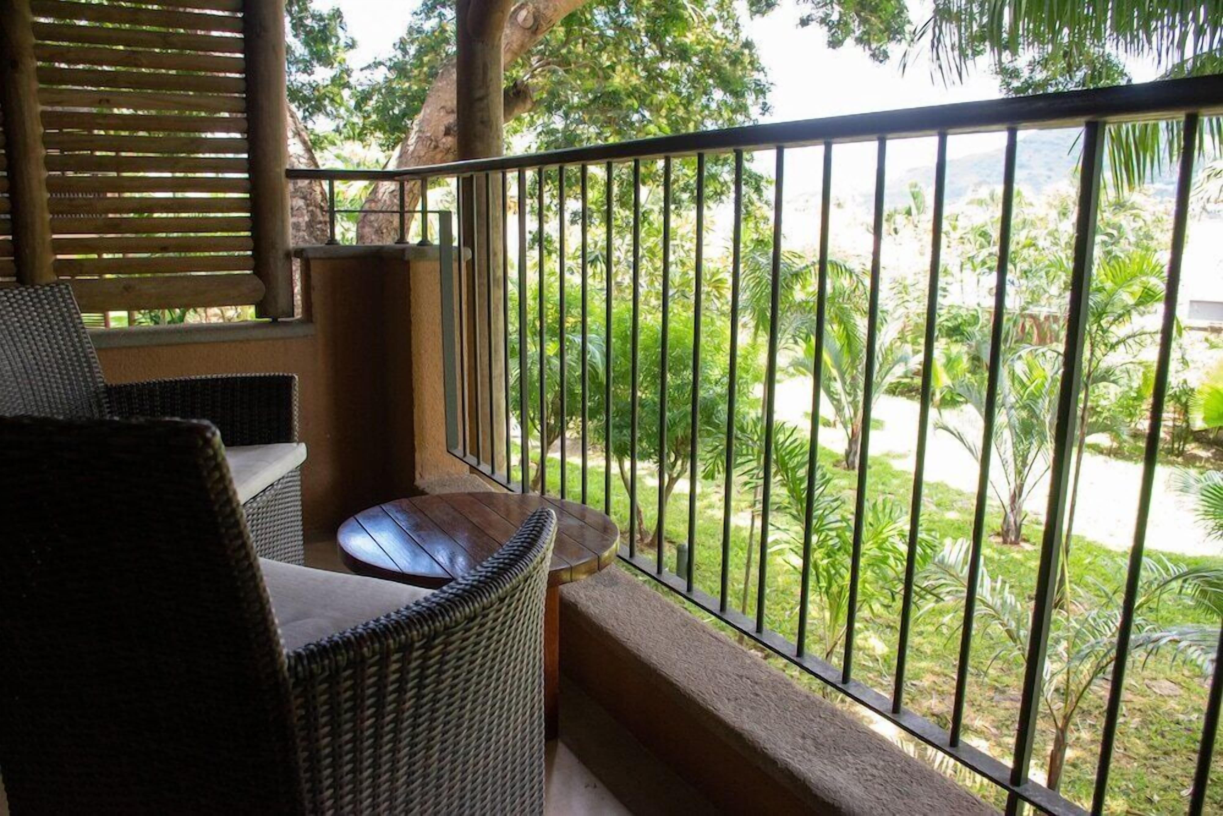 Uma varanda com móveis de vime, uma mesa redonda pequena e vista para um jardim exuberante.