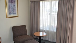 Um quarto com uma poltrona marrom, uma mesa redonda pequena, uma obra de arte emoldurada na parede e uma janela grande com cortinas.