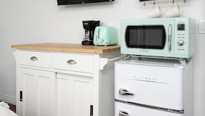 Uma cozinha compacta com micro-ondas, torradeira e cafeteira, tudo sobre um armário branco, uma TV fixada na parede e uma prateleira com chávenas.
