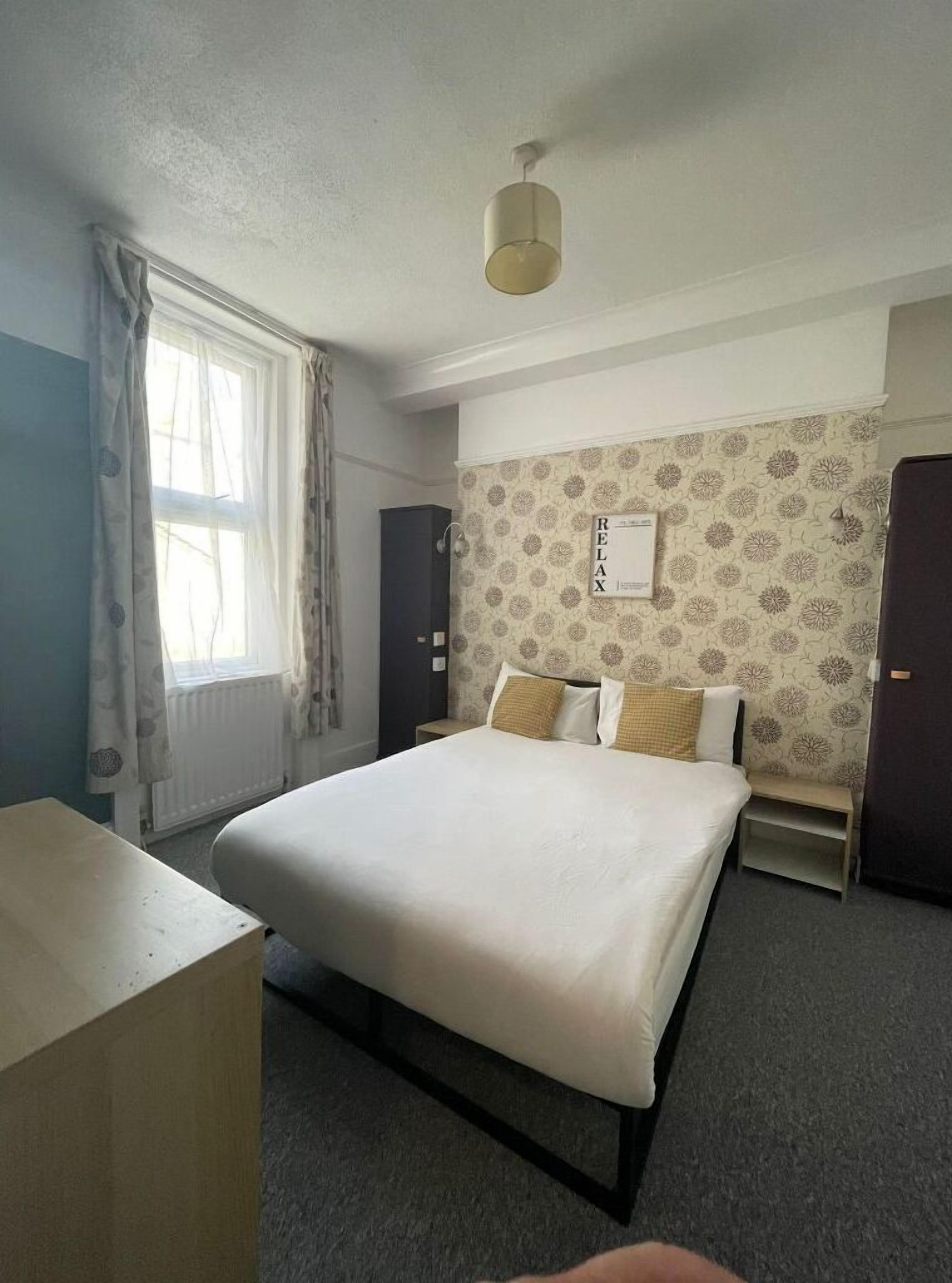 Quarto com cama, mesa de cabeceira, guarda-roupa, janela com cortinas e parede com padrão floral.