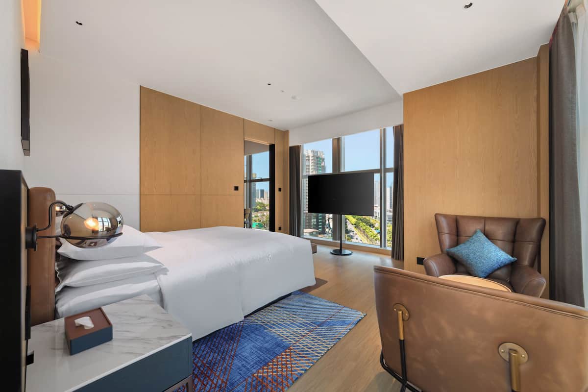 Quarto de hotel moderno com uma cama grande, uma televisão de tela plana, uma poltrona de couro e vista para a cidade por grandes janelas.
