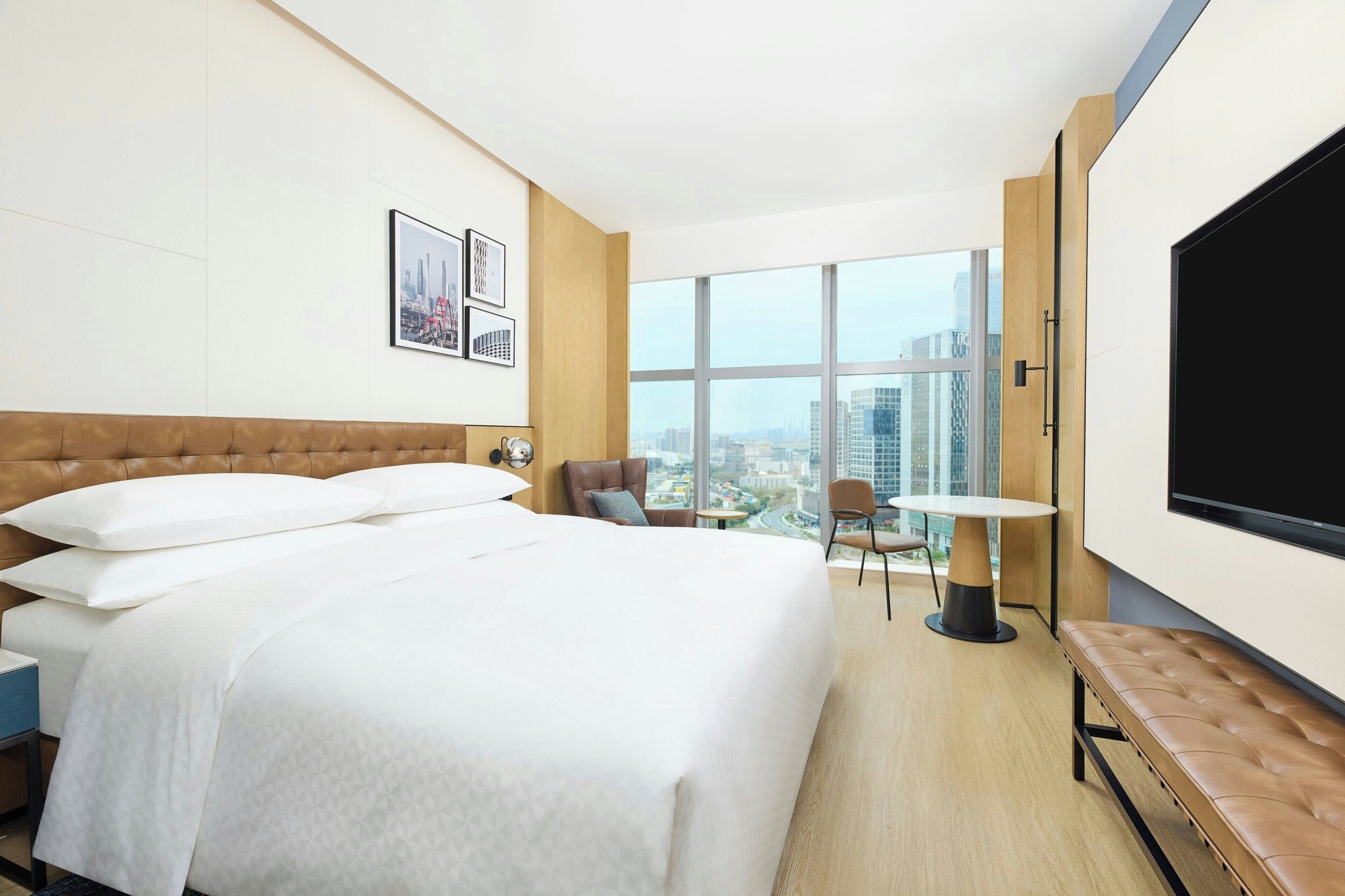 Quarto de hotel com uma cama grande, uma escrivaninha, uma cadeira, uma mesa redonda, uma televisão e vista para a cidade.