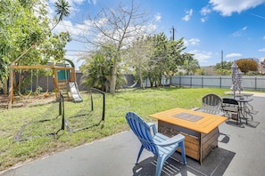 Um quintal com uma mesa de madeira, cadeiras azuis, uma rede e um parquinho.
