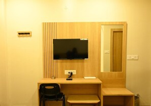 Quarto de hotel com escrivaninha de madeira, televisão na parede, uma cadeira e uma pequena prateleira.
