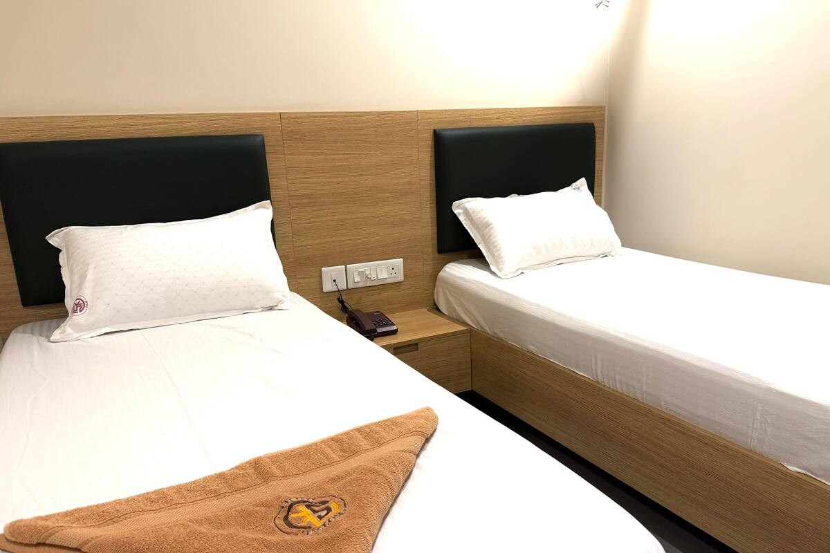 Quarto de hotel com duas camas, cada uma com colcha e travesseiro brancos, painel de cabeceira de madeira, mesa de cabeceira com telefone e toalha marrom no chão.
