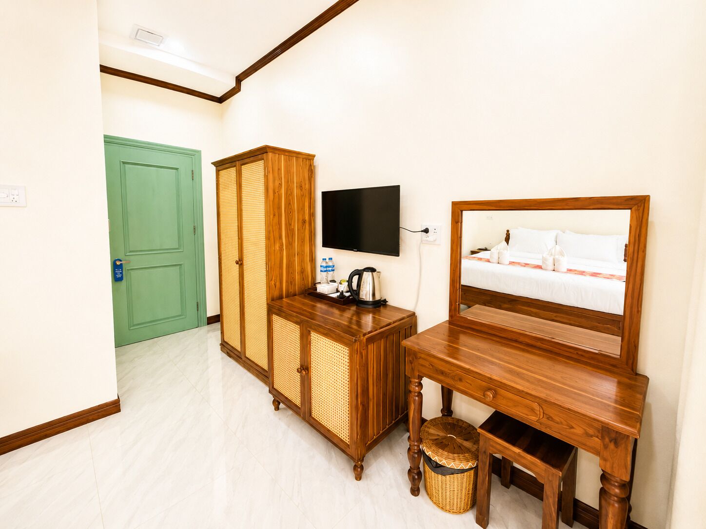 Quarto de hotel com uma cama, televisão, armário de madeira, cesto de vime e uma porta verde.