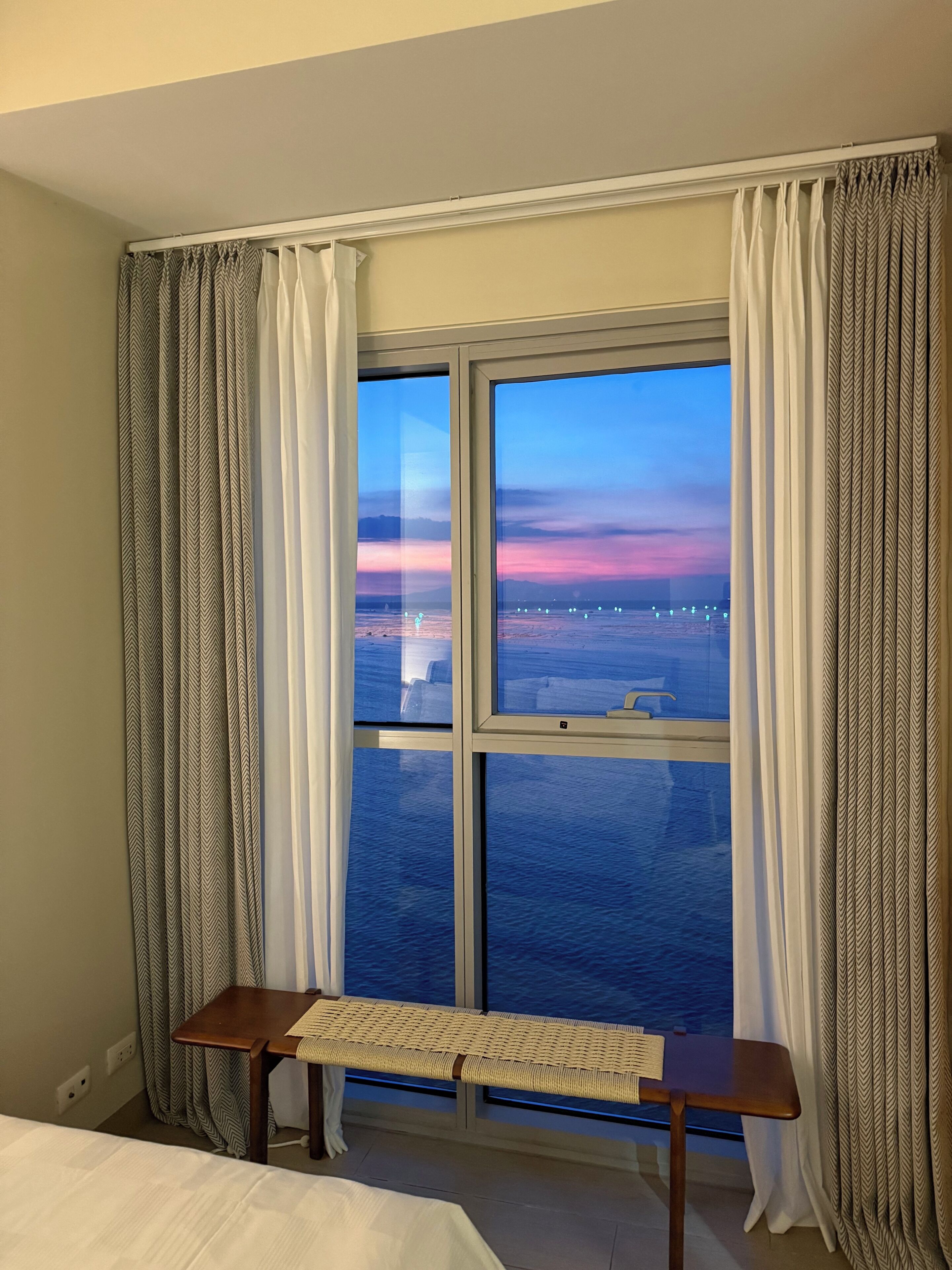 Quarto de hotel com uma janela grande, uma cama, uma mesinha e cortinas.