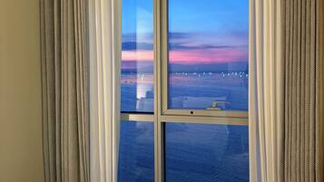 Quarto de hotel com uma janela grande, uma cama, uma mesinha e cortinas.