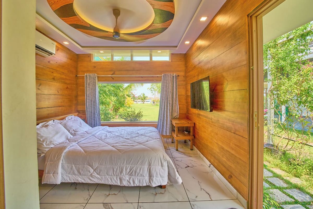 Quarto com cama, paredes de madeira, ventilador de teto e vista para o verde lá fora.