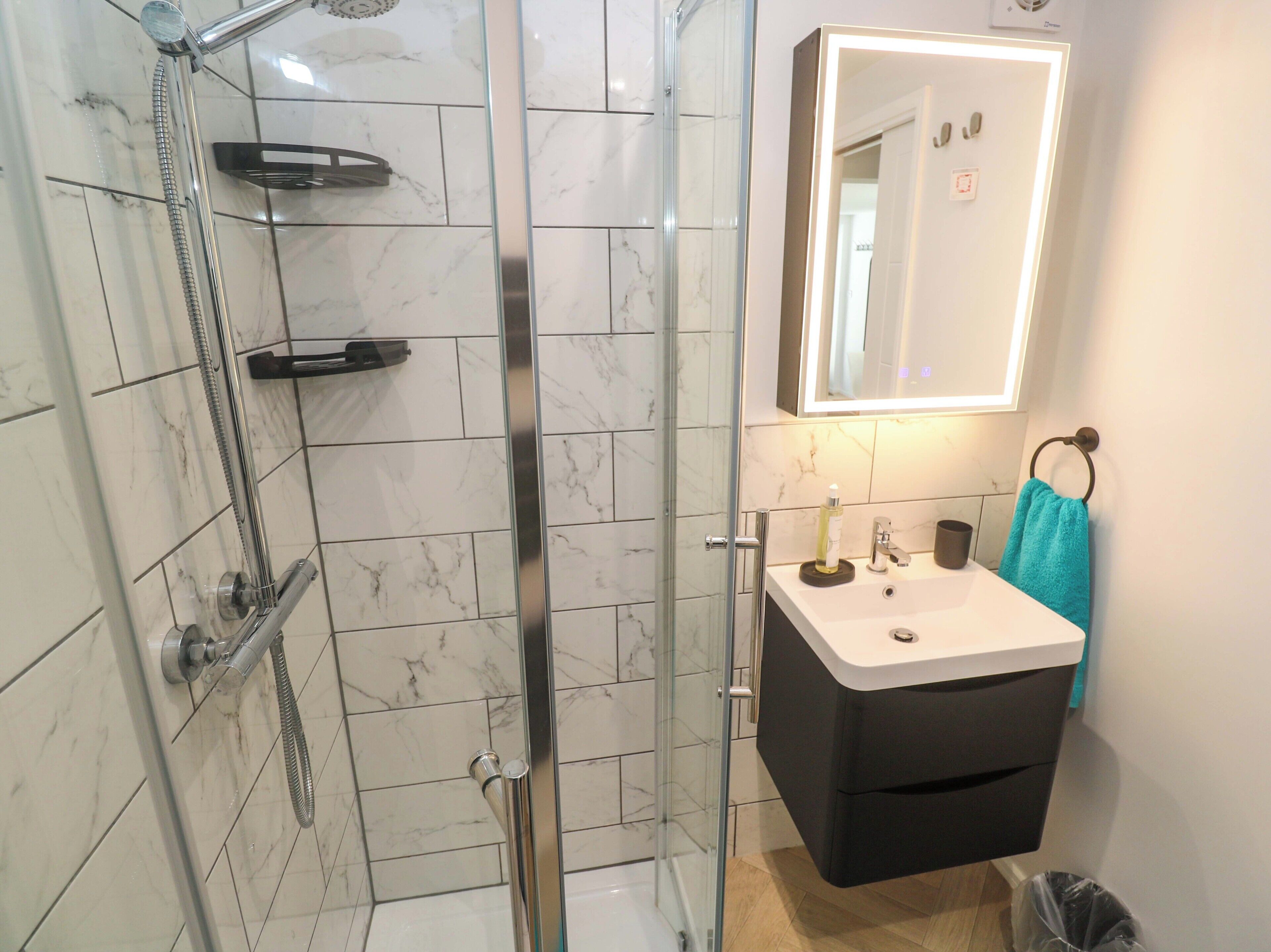 Casa de banho com chuveiro de vidro, pia branca com armário preto, espelho com luz e toalha azul pendurada num suporte.