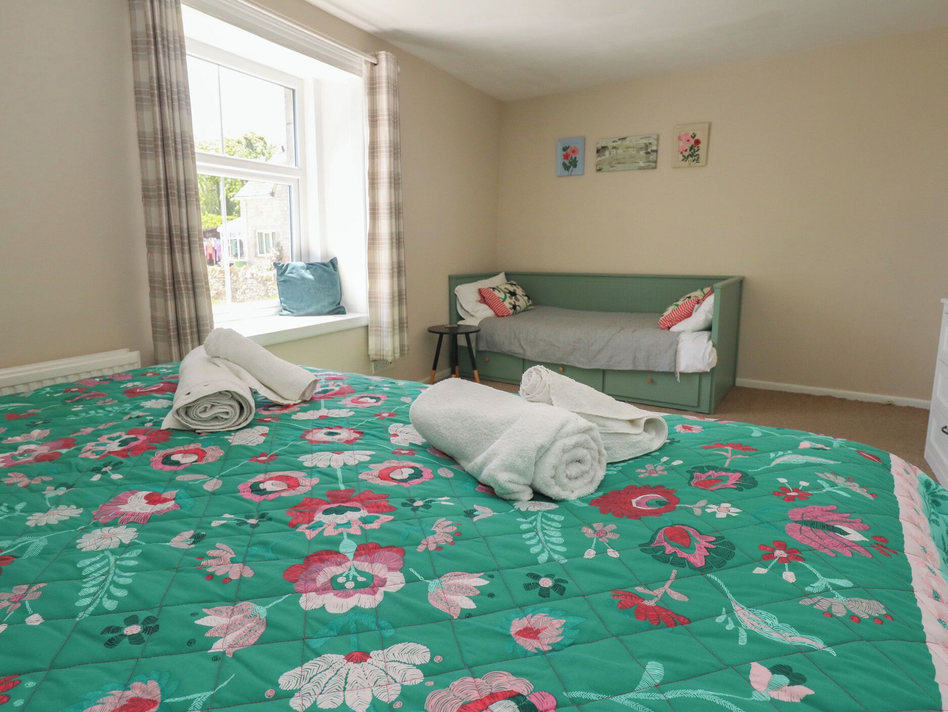 Quarto com colcha de cama estampada em verde, duas camas de solteiro, uma janela com cortinas e uma mesa pequena.