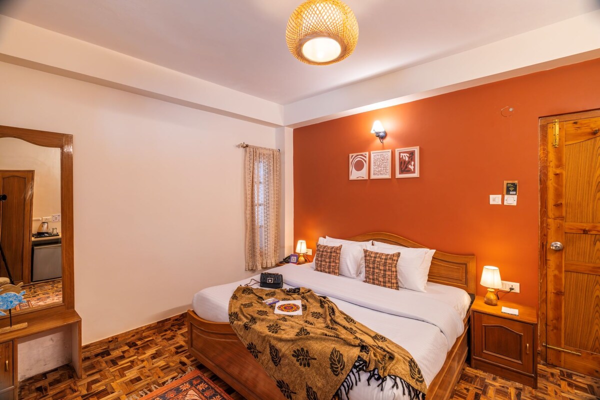 Quarto de hotel com cama de madeira, mesa de cabeceira, luminária na parede e porta com painel de madeira.