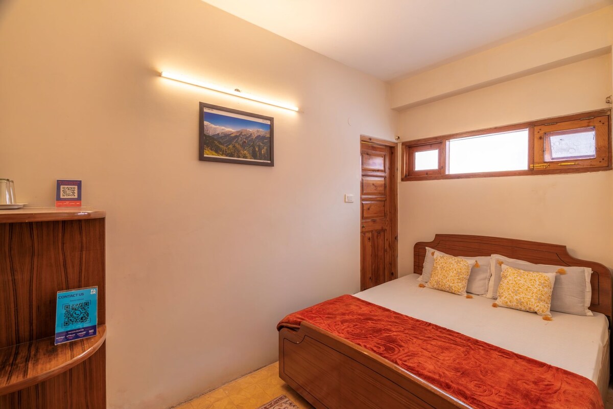 Quarto com cama de madeira, mesa de cabeceira, prateleira na parede, quadro emoldurado e janela com persianas de madeira.