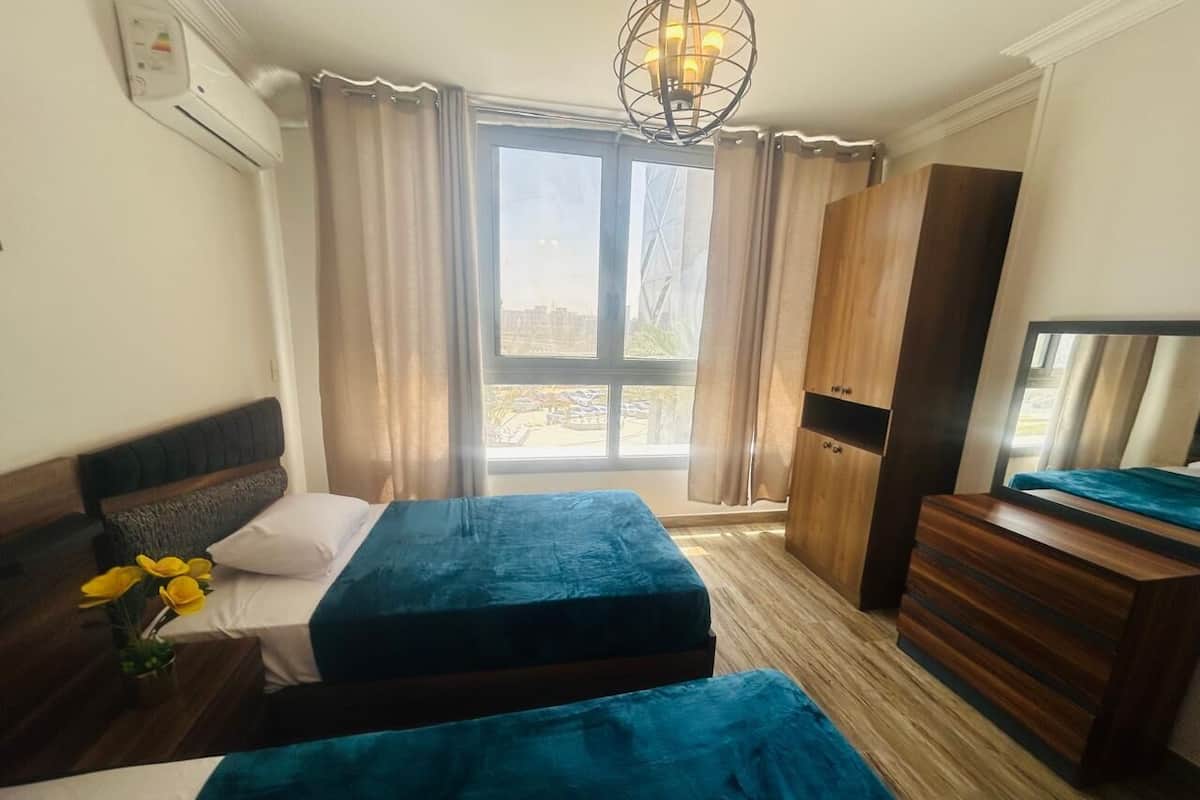Quarto de hotel com duas camas, um guarda-roupa de madeira, uma escrivaninha com espelho e uma janela com cortinas.