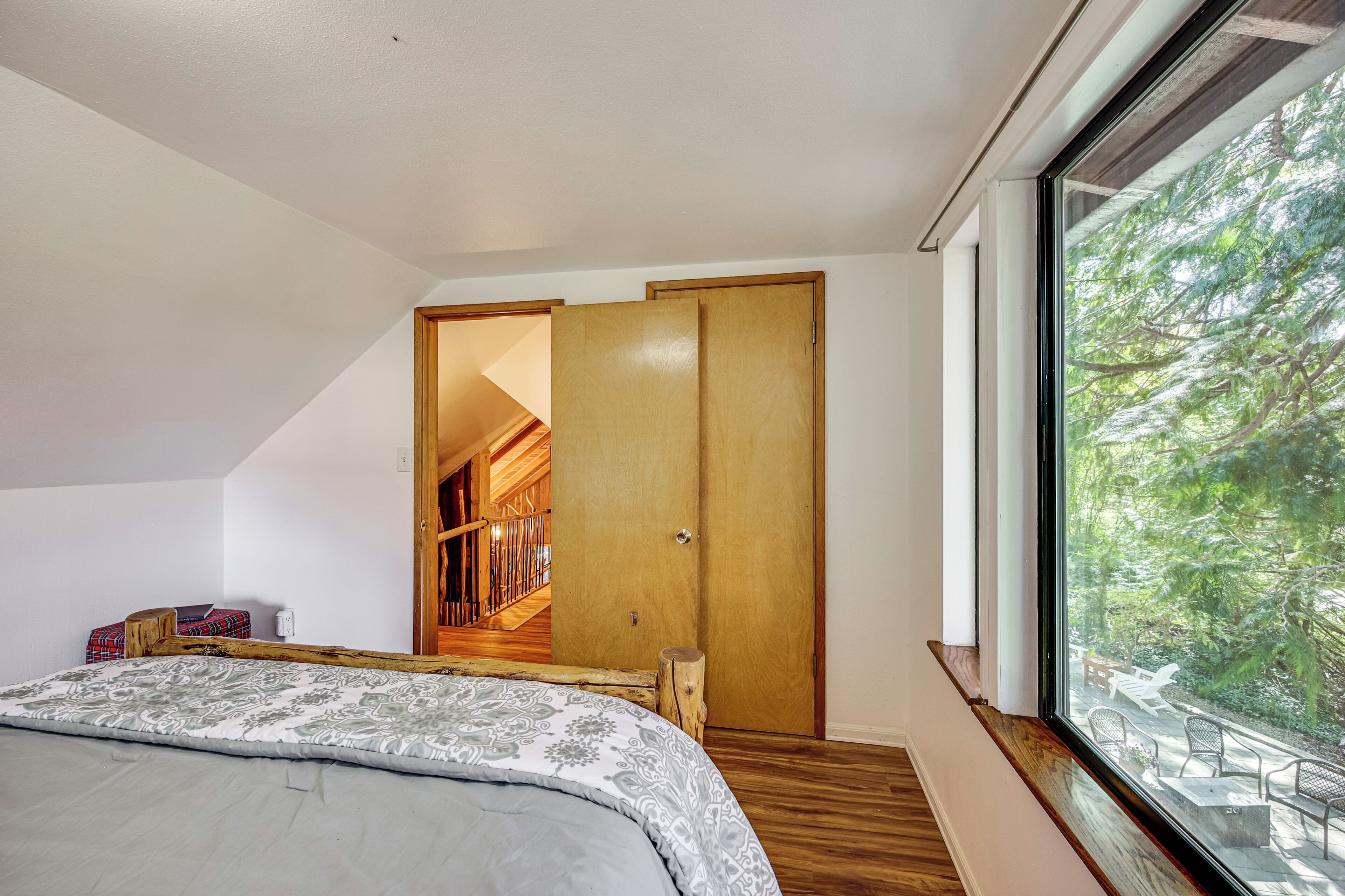 Quarto com cama, guarda-roupa de madeira, janela com vista para árvores e escada que dá acesso a um mezanino.