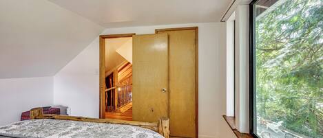 Quarto com cama, guarda-roupa de madeira, janela com vista para árvores e escada que dá acesso a um mezanino.