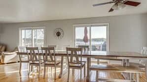 Uma área de refeições com uma mesa e cadeiras de madeira, um relógio na parede e vista através de grandes janelas.