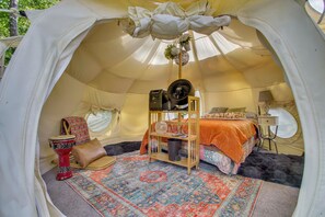 Uma tenda com uma cama, uma mesa pequena, uma cadeira, um ventilador e um tapete.