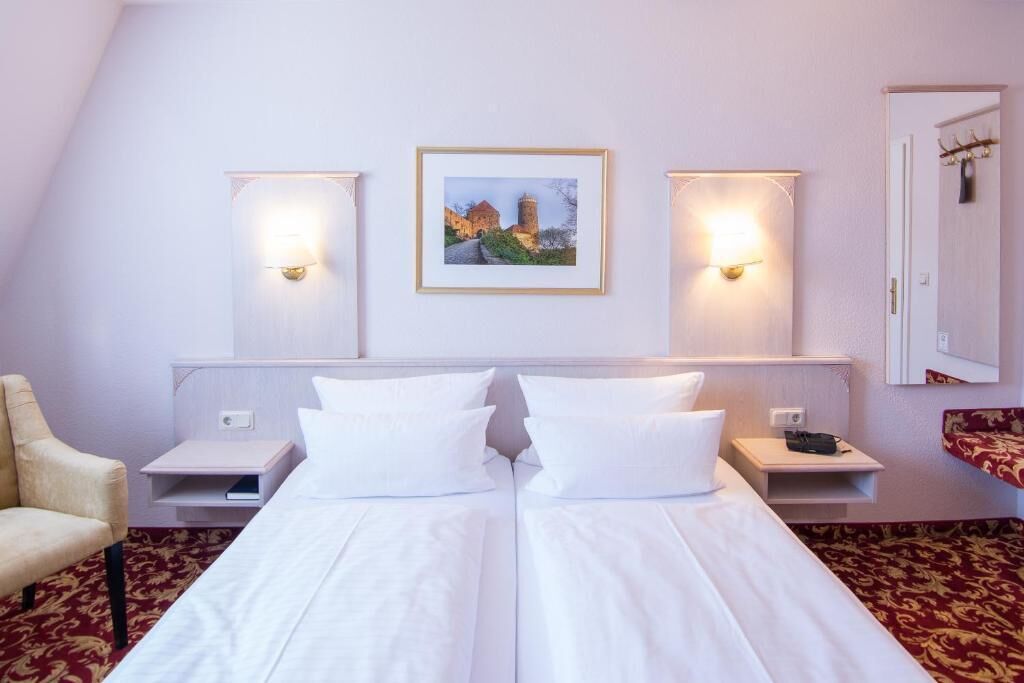 Quarto de hotel com duas camas, mesas de cabeceira, uma cadeira, um quadro emoldurado na parede e uma porta com um cabide.
