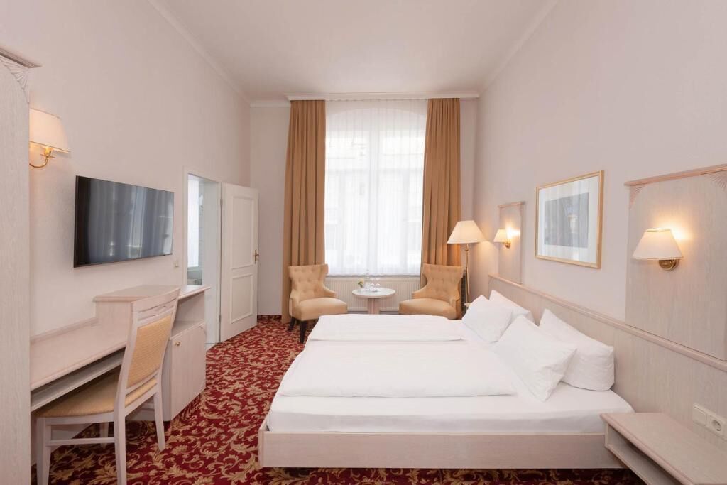 Quarto de hotel com cama, escrivaninha, cadeiras, televisão e janela com cortinas.