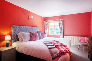 Quarto com cama, mesa de cabeceira, penteadeira, janela com cortinas e luminária fixada na parede.
