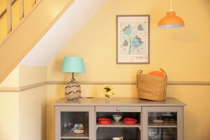 Um canto com um armário cinza, uma luminária azul, um vaso com flores amarelas, uma cesta de vime e um quadro na parede.