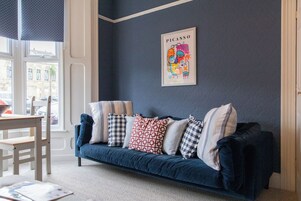 Um quarto com um sofá azul escuro, almofadas estampadas, uma obra de arte emoldurada na parede e uma janela com vista para a rua.
