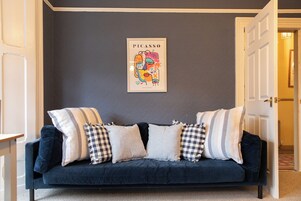 Um sofá azul escuro com almofadas listradas e quadriculadas, uma obra de arte emoldurada na parede e uma porta que dá para outro quarto.