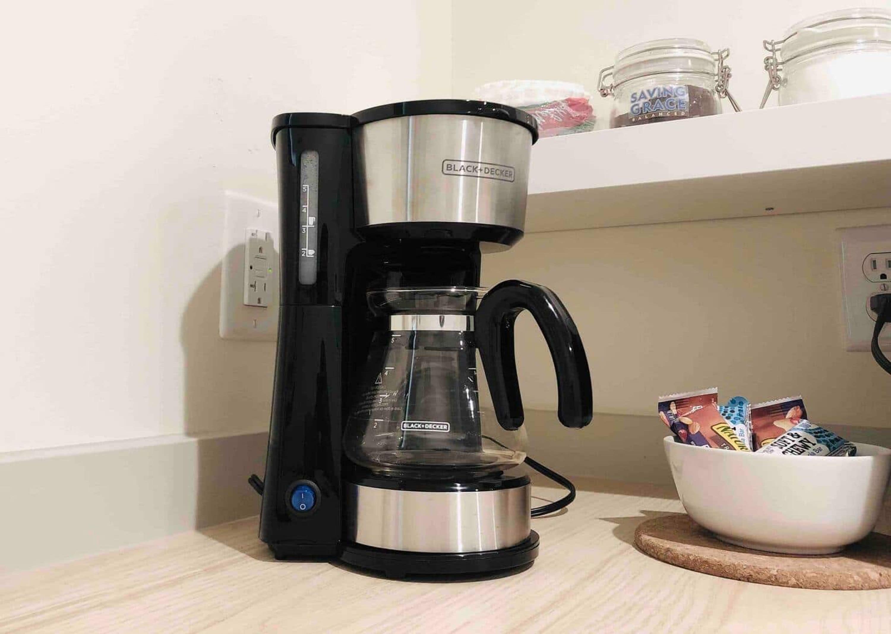 Uma cafeteira BLACK+DECKER sobre uma bancada de cozinha, com uma tigela branca contendo cápsulas de café e um frasco rotulado 'SAVING GRACE BALANCE'.