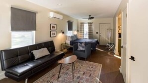 Uma sala moderna com sofá, mesinha, tapete estampado, cama com cabeceira e janela com persianas.