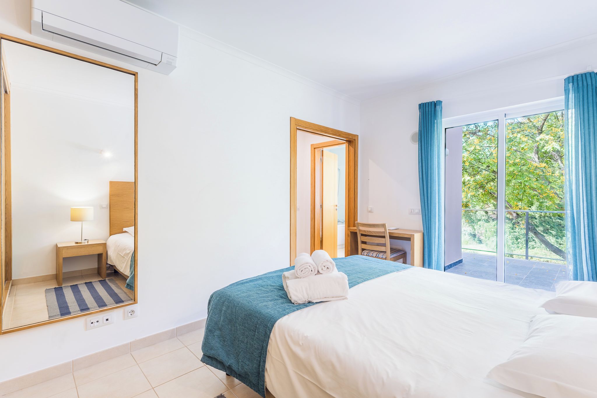 Quarto de hotel com cama, escrivaninha, cadeira, espelho e varanda com cortinas.
