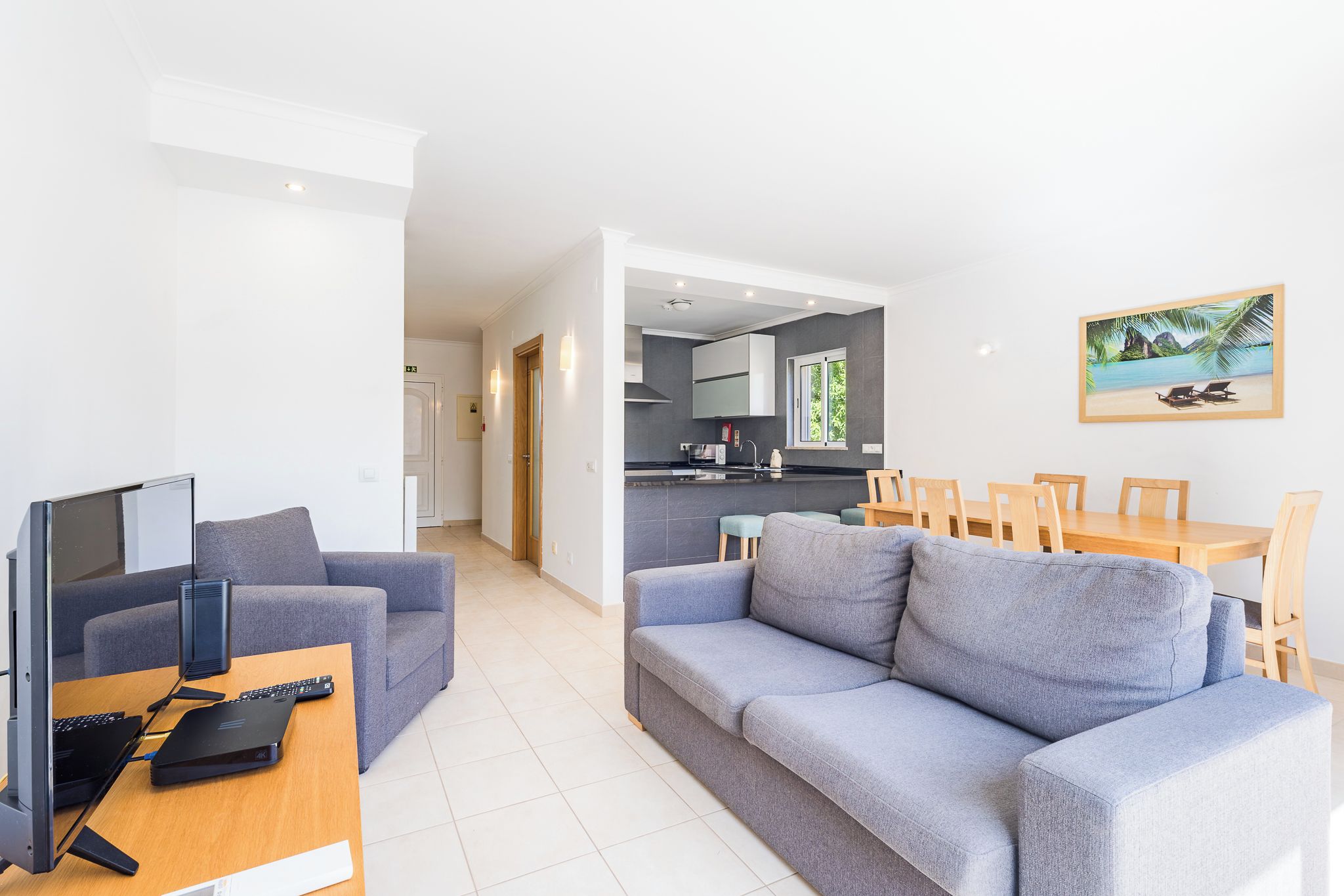 Uma sala de estar moderna com um sofá cinzento, uma mesa de centro de madeira, uma TV e um laptop. Há uma área de jantar com uma mesa e cadeiras de madeira.