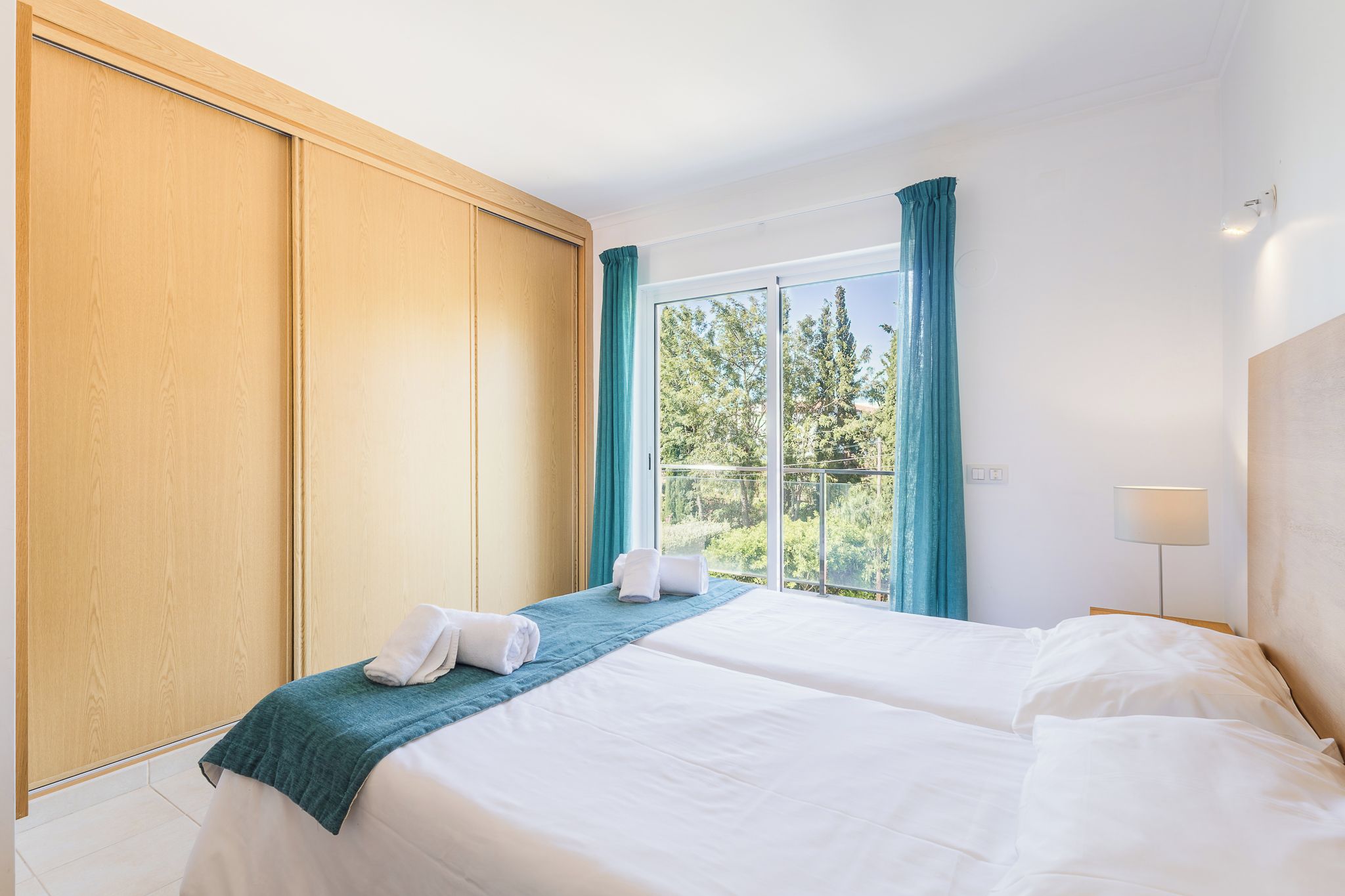 Quarto de hotel com uma cama grande, um guarda-roupa de madeira, uma janela com cortinas e uma mesa de cabeceira com abajur.