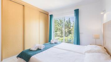 Quarto de hotel com uma cama grande, um guarda-roupa de madeira, uma janela com cortinas e uma mesa de cabeceira com abajur.