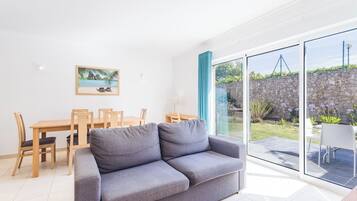 Uma sala de estar moderna com um sofá cinza, mesa de jantar de madeira, cadeiras e uma grande porta de vidro deslizante que dá para uma área externa com parede de pedra e plantas.