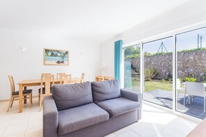 Uma sala de estar moderna com um sofá cinza, mesa de jantar de madeira, cadeiras e uma grande porta de vidro deslizante que dá para uma área externa com parede de pedra e plantas.