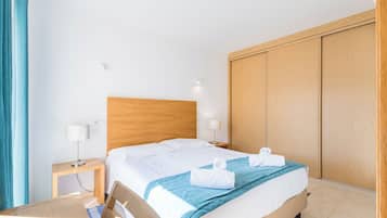 Quarto de hotel com cabeceira de madeira, cama com roupa de cama branca e manta azul, além de um guarda-roupa de madeira.