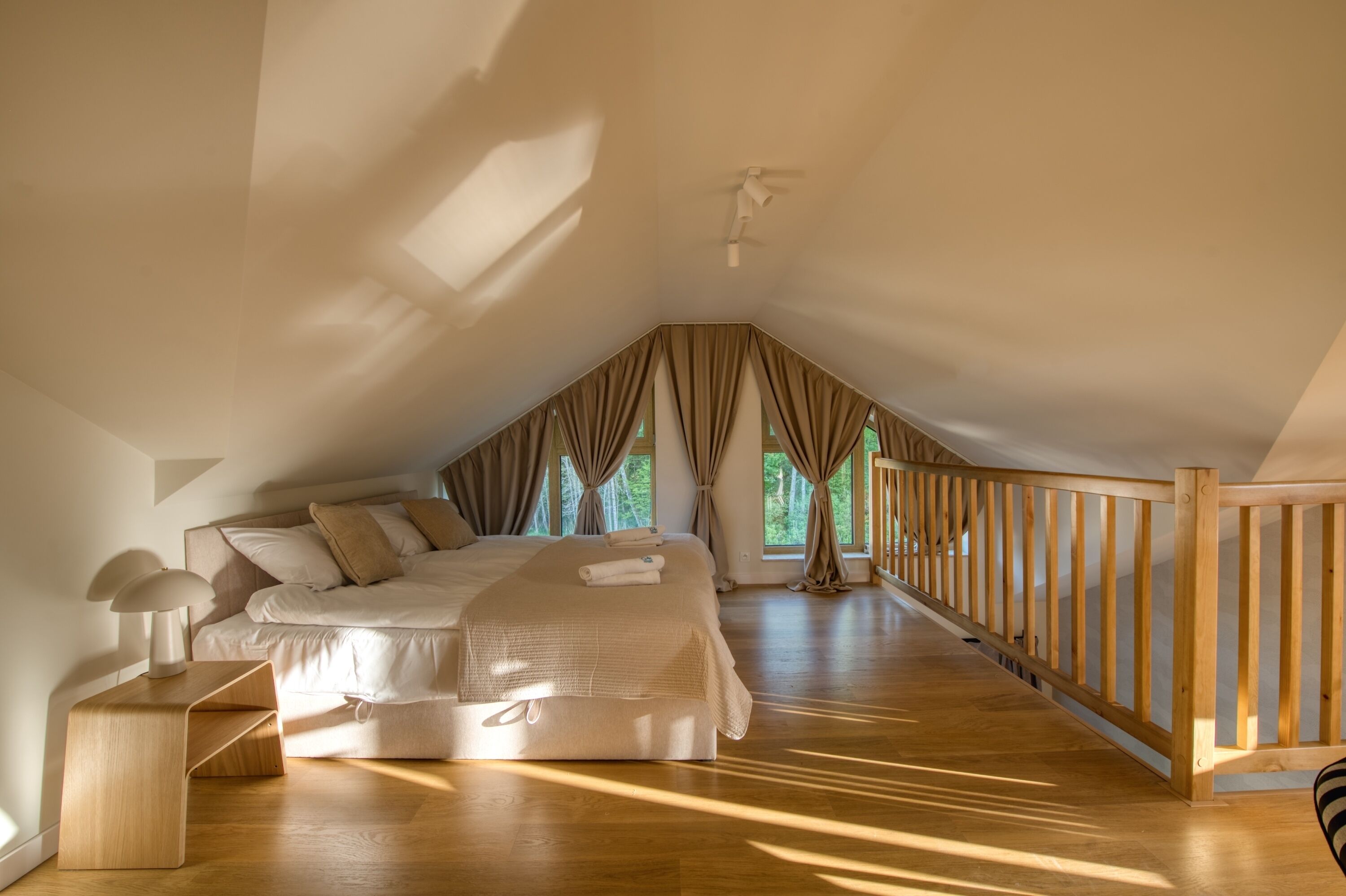 Quarto com teto inclinado, cama, mesa de cabeceira e corrimão de madeira.