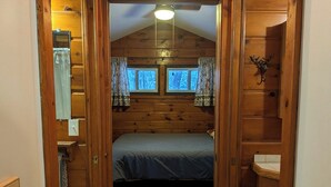 Interior de uma cabana de madeira com uma cama, duas janelas com cortinas, uma luminária no teto e um aplique na parede.