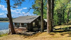 Uma cabana com varanda, localizada à beira de um lago, rodeada por árvores.