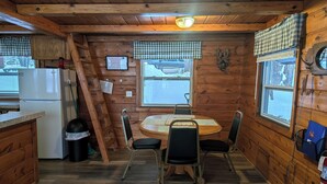Interior de uma cabana de madeira com uma mesa de jantar, cadeiras, um refrigerador, uma escada e uma janela com vista para uma paisagem nevada.