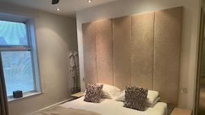 Quarto com uma cama grande, cabeceira, janela e mesa de cabeceira.