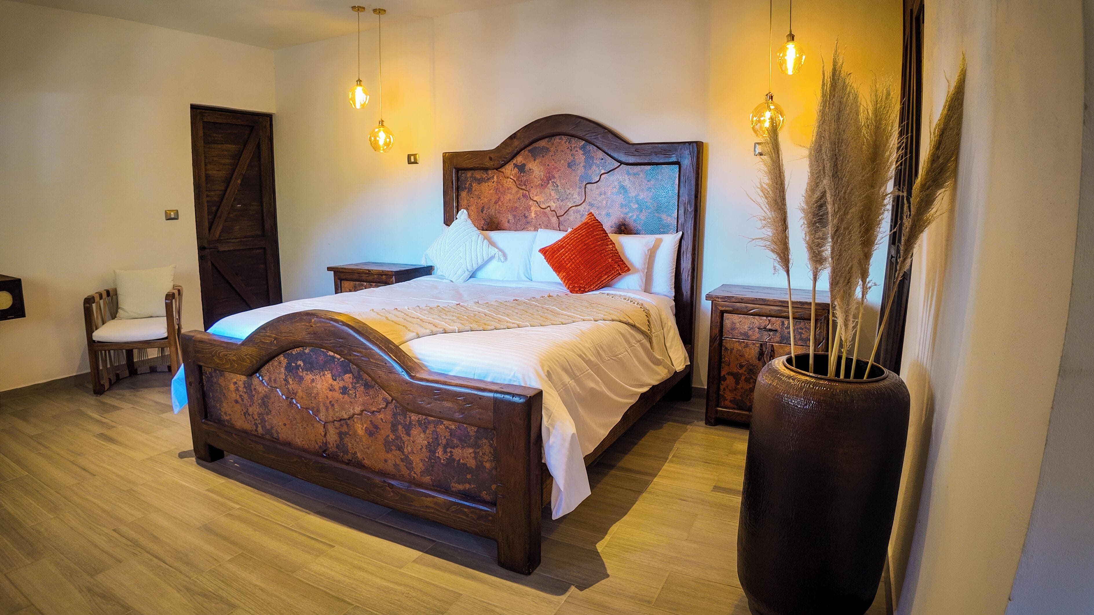 Quarto com cama de madeira, mesa de cabeceira, cadeira e um vaso grande com capim-do-campo.