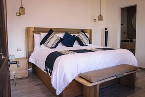 Cama bem arrumada com cabeceira de madeira, mesa de cabeceira com abajur e um banco no rodapé da cama.