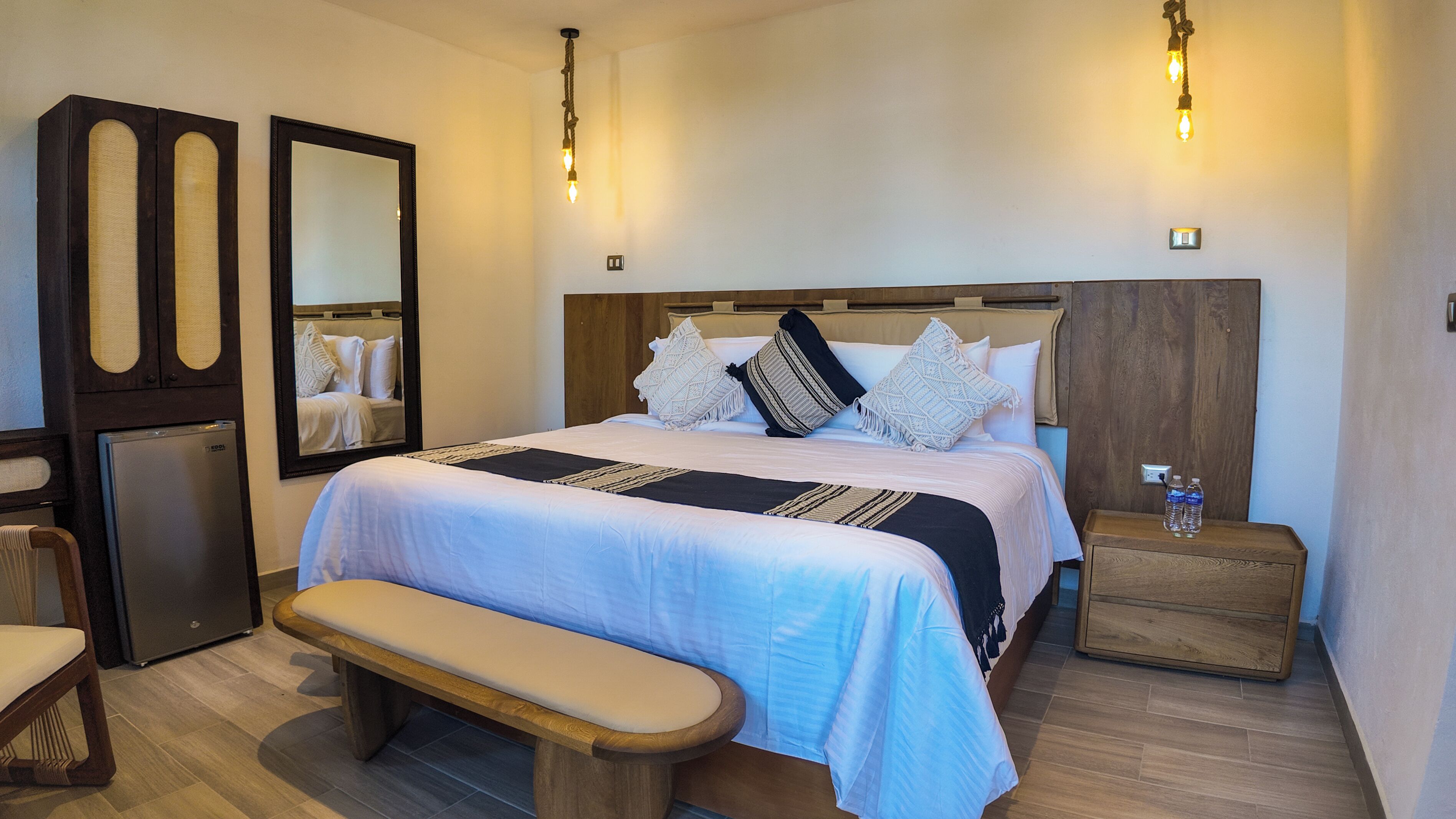 Quarto bem arrumado com cama, um banco, mesas de cabeceira, um espelho e um frigorífico.