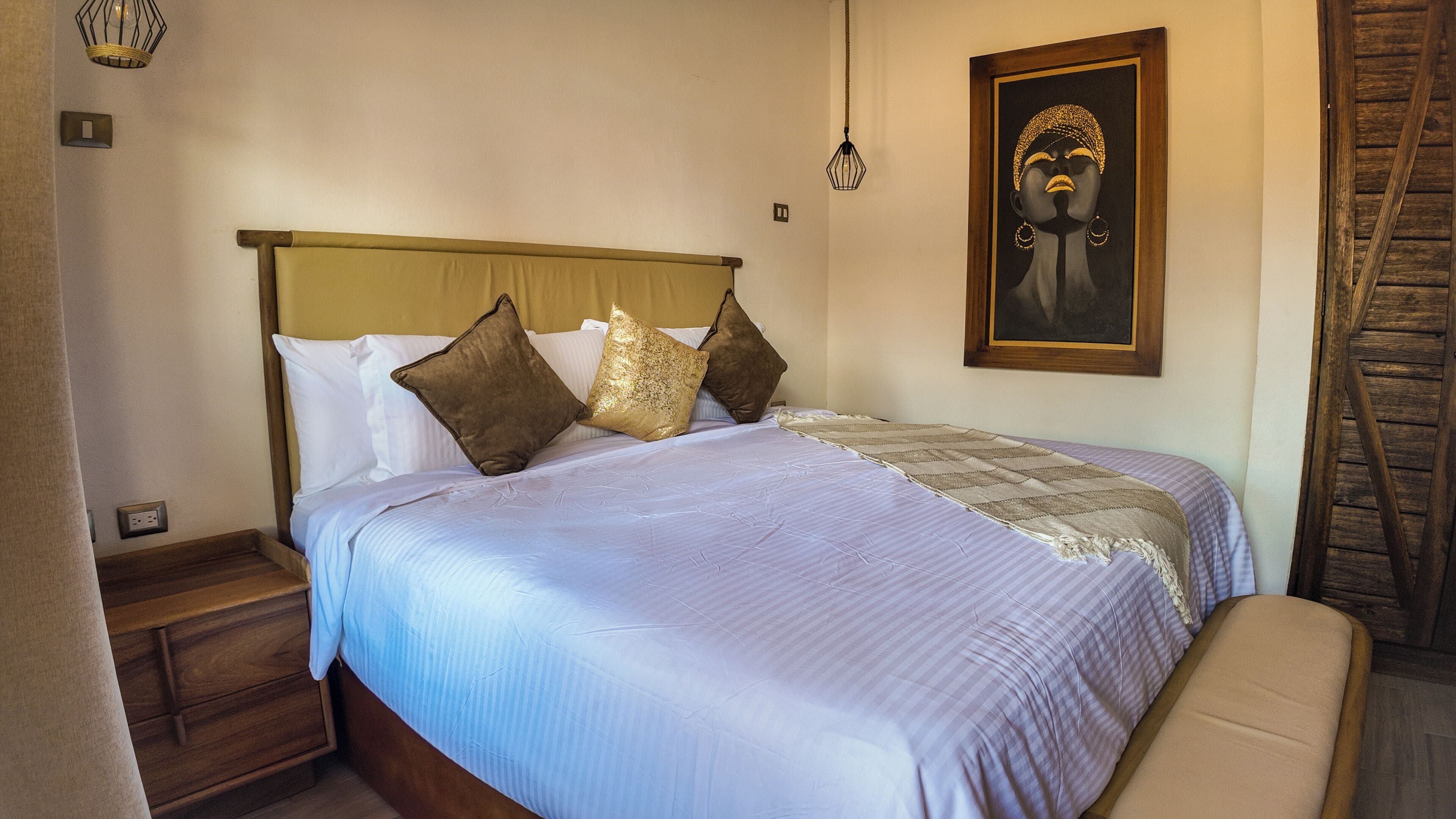Cama bem arrumada com cabeceira, mesa de cabeceira e um banquinho.