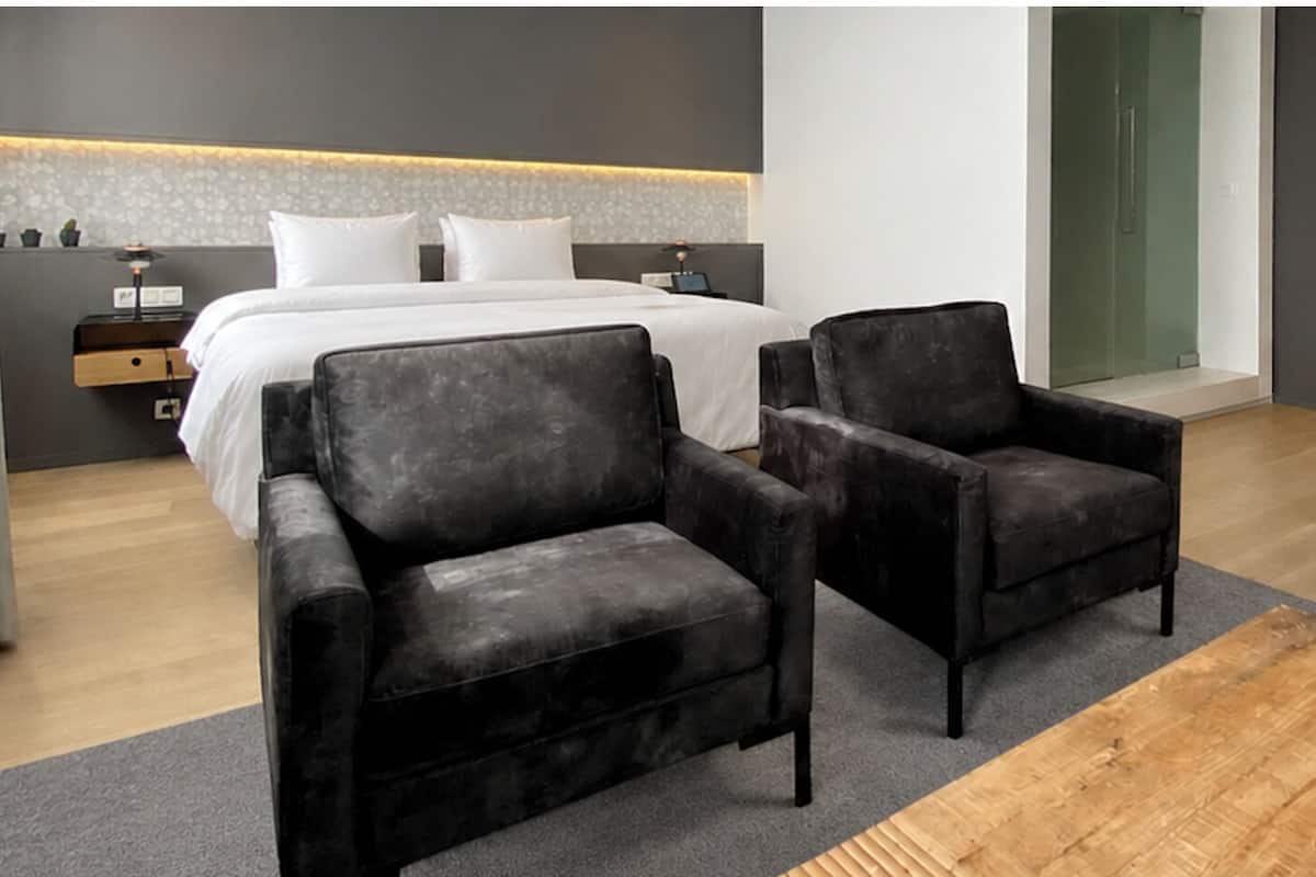 Quarto de hotel moderno com uma cama, duas poltronas, uma mesa de centro de madeira, uma mesinha de cabeceira e uma luminária de parede.